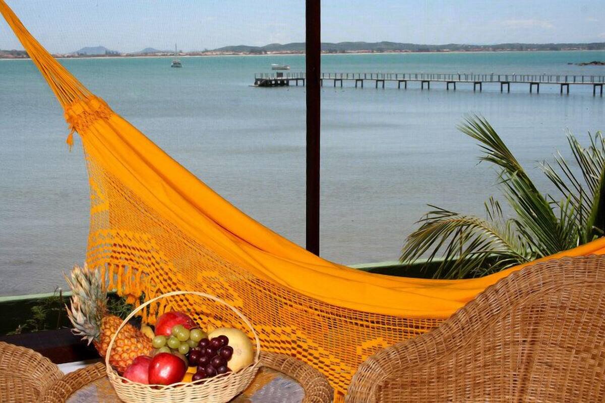 Uma cadeira rede com uma cesta de vime contendo frutas e uma abacaxi, com vista para um mar tranquilo e um cais ao longe.