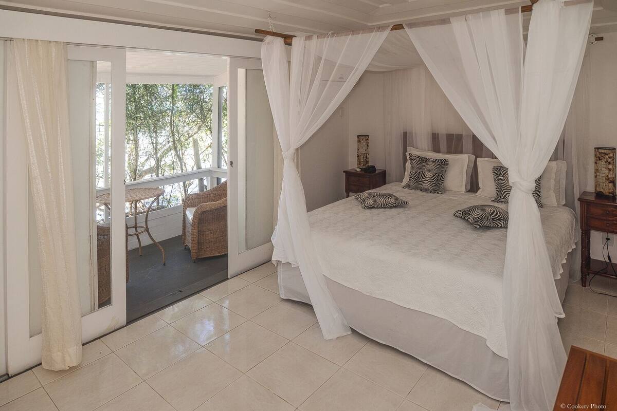 Quarto com cama de dossel, varanda com móveis de vime e vista para árvores.