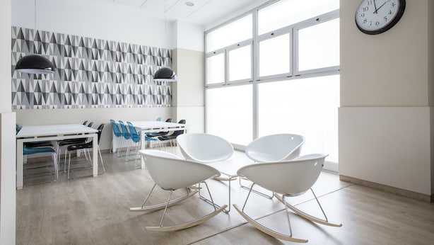 Uma sala de reuniões moderna com uma mesa branca, cadeiras azuis, um design geométrico na parede e uma janela grande.