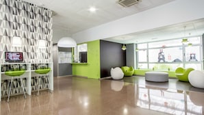 Uma área de recepção moderna com uma parede de padrão geométrico, um balcão de recepção com tela digital e uma área de estar com cadeiras verdes e brancas.