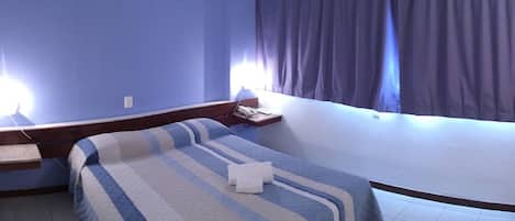 Quarto de hotel com cama, mesa de cabeceira, luminária e janela com cortinas.