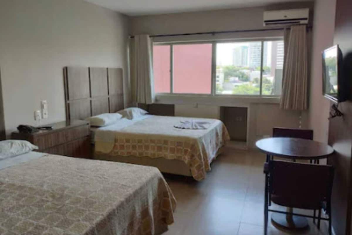 Quarto de hotel com duas camas, uma mesinha e uma janela com vista para prédios.