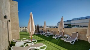 Uma área de lounge no terraço com espreguiçadeiras e guarda-sóis, grama sintética verde e vista para o horizonte da cidade.