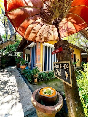 Um sinalizador com a inscrição 'Room 101-109 Pool' e um ambiente externo rústico, com uma árvore, plantas em vasos e um edifício tradicional.