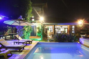 Área de piscina com espreguiçadeiras e mesa, cercada por plantas e um edifício com entrada visível.