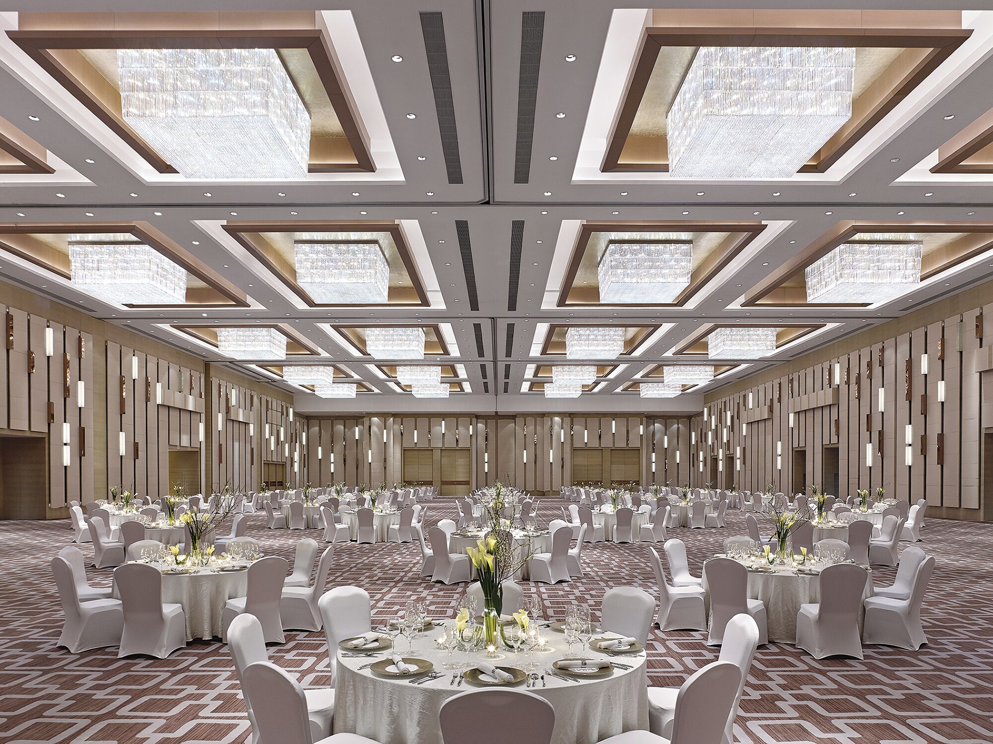 Uma ampla sala de banquetes com mesas redondas, cadeiras brancas e lustres.