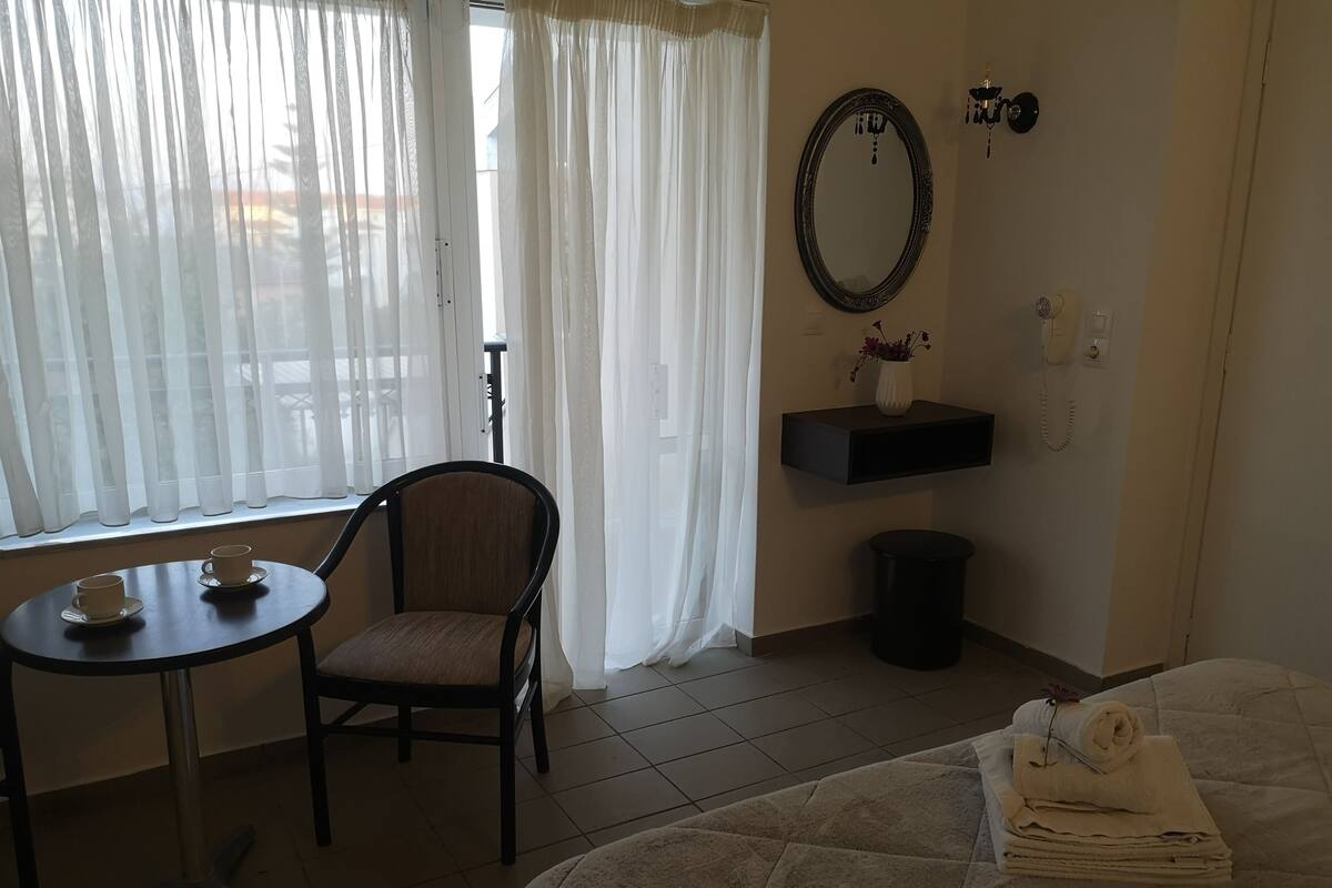 Quarto de hotel com uma cama, uma cadeira, uma mesinha com chávenas, um espelho, uma estante com um vaso e um cesto de lixo.
