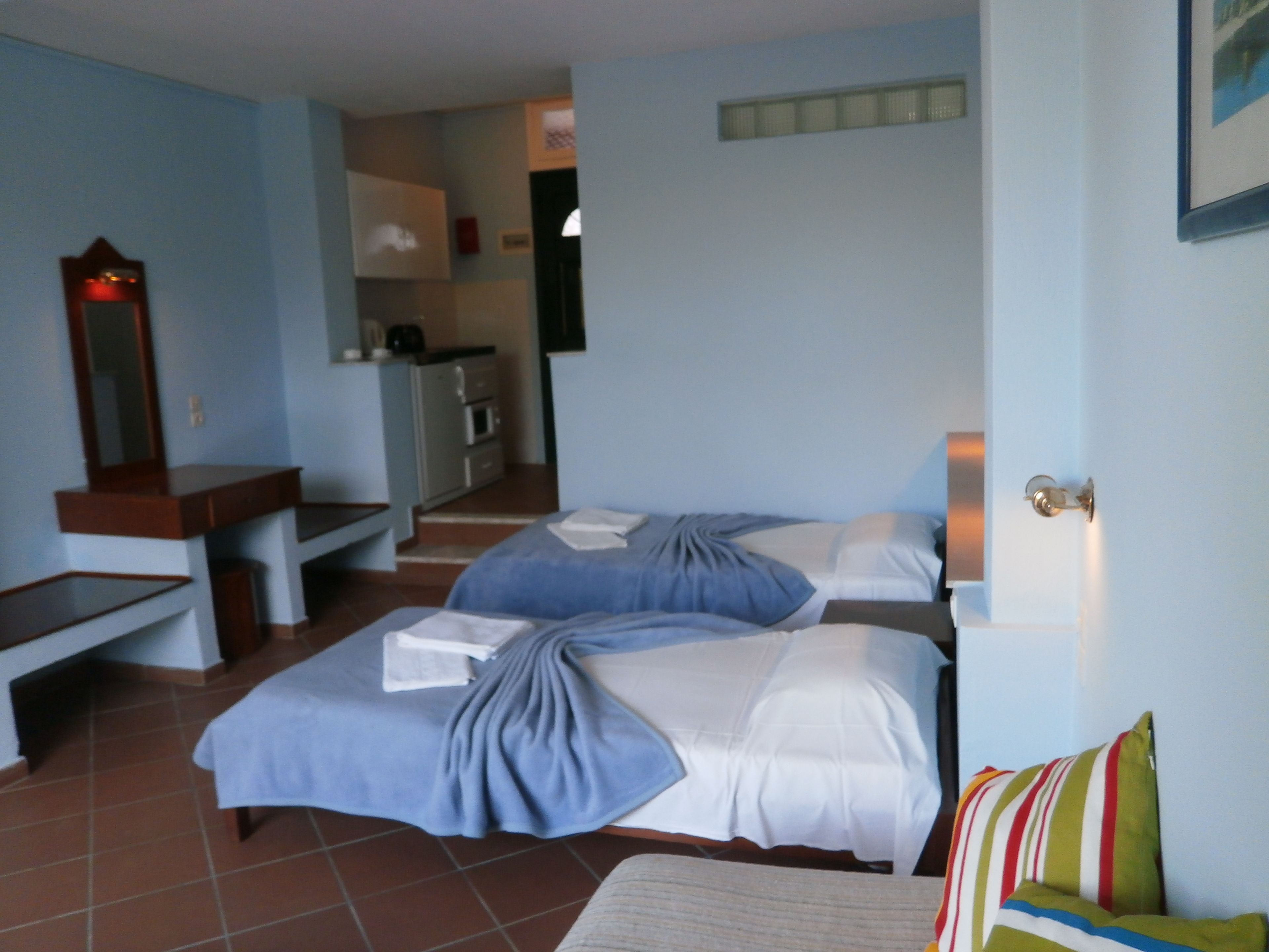 Quarto de hotel com duas camas, uma escrivaninha, um espelho, cozinha compacta e banheiro.