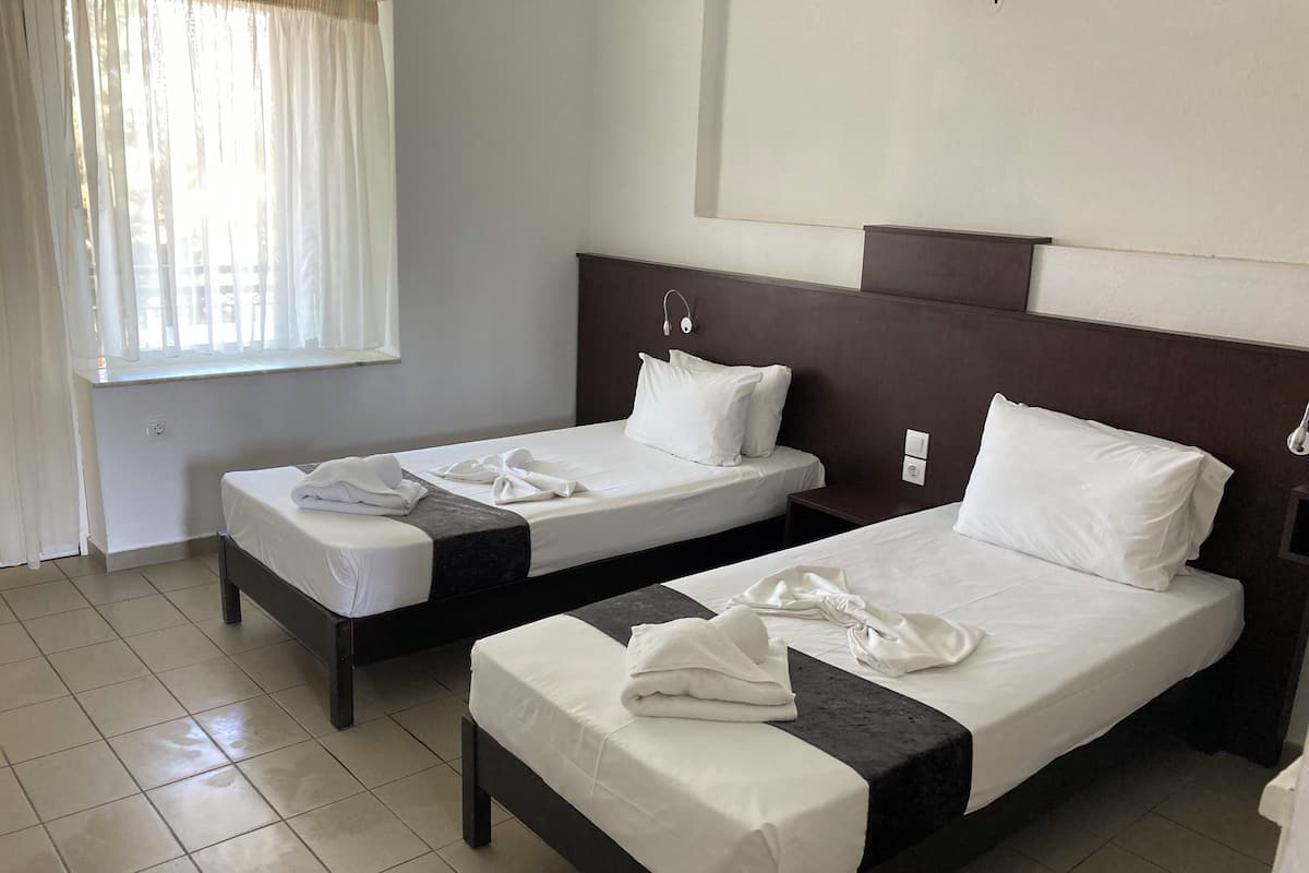 Quarto de hotel com duas camas de solteiro, uma janela grande com cortinas e chão em azulejos.