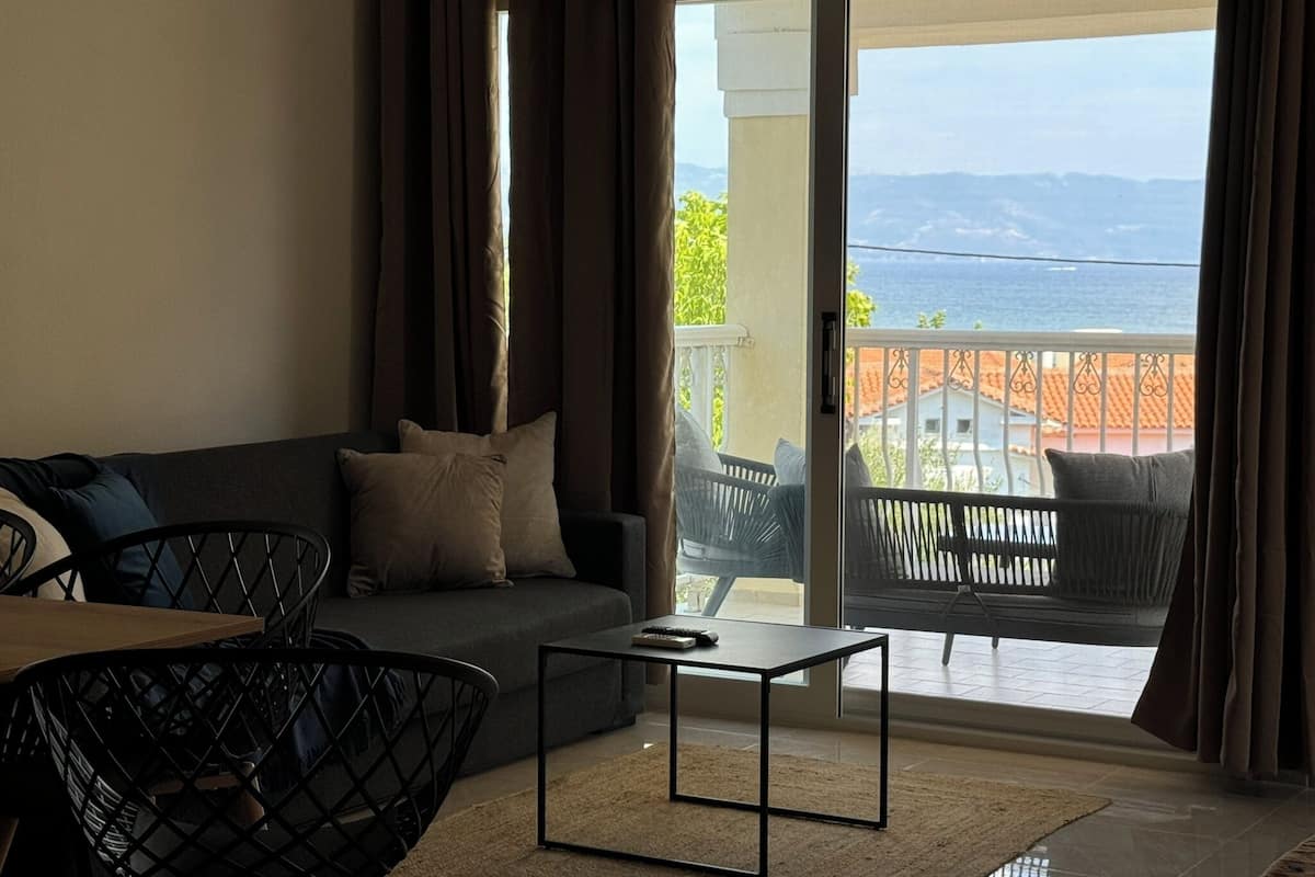 Uma sala de estar com sofá, mesa de centro e vista para o mar através de uma porta de vidro deslizante.
