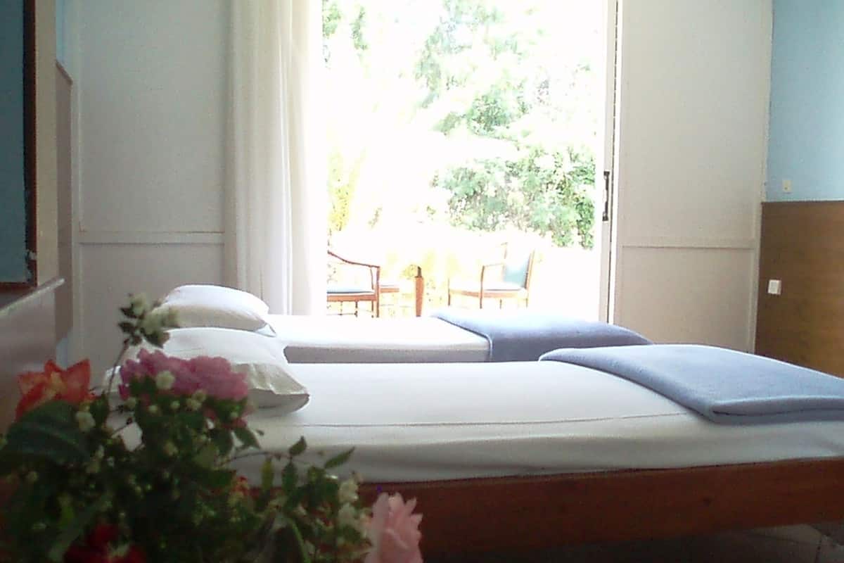 Quarto com cama, janela e vista para área externa com cadeiras e vegetação.