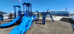 Um parquinho com escorregadores e estruturas de escalada, cercado por uma área pavimentada e um edifício com placas.