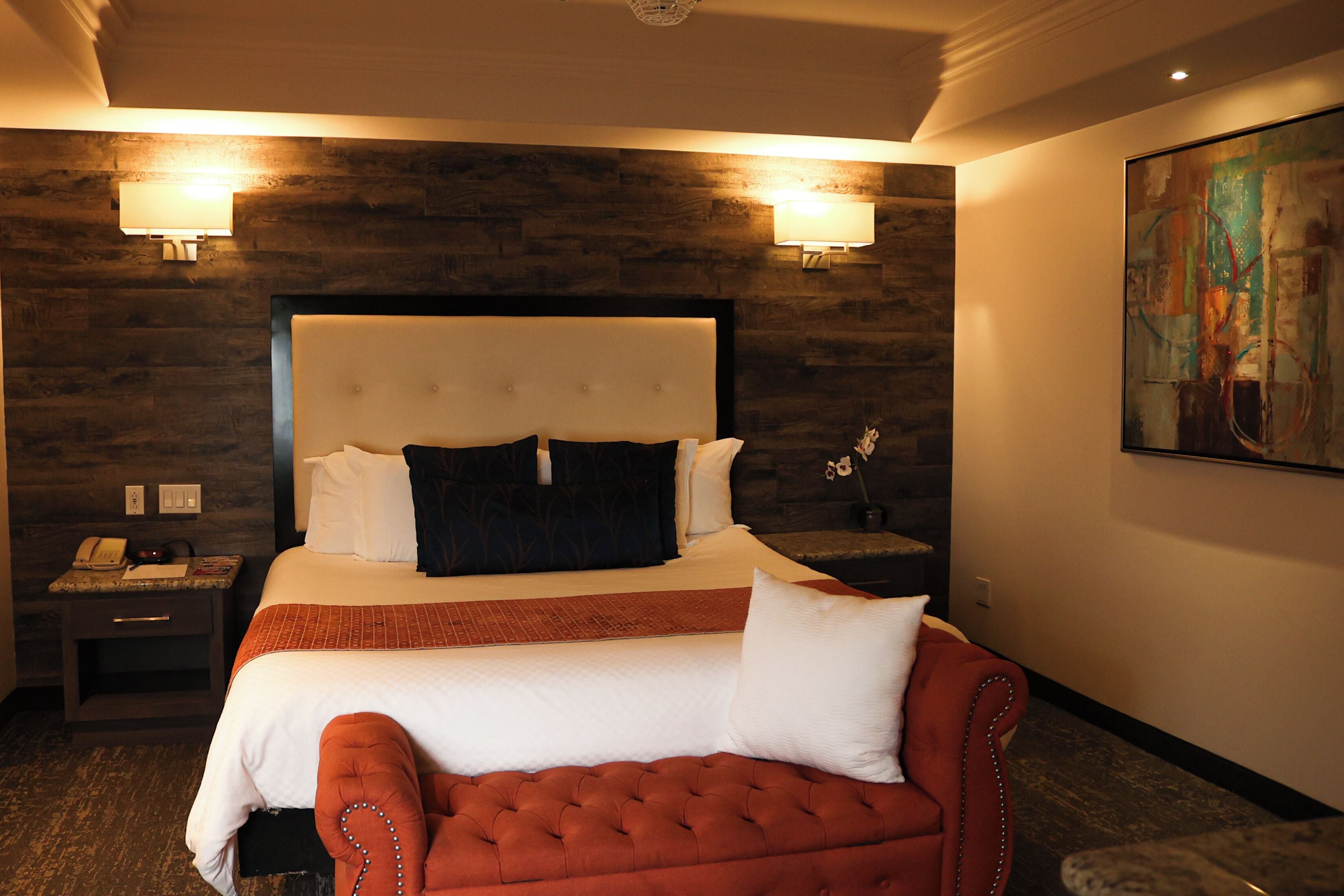 Quarto de hotel com uma cama grande, uma poltrona estofada vermelha, duas mesas de cabeceira com luminárias e um quadro na parede.
