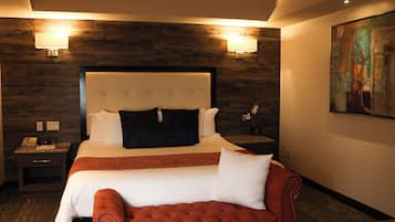 Quarto de hotel com uma cama grande, uma poltrona estofada vermelha, duas mesas de cabeceira com luminárias e um quadro na parede.