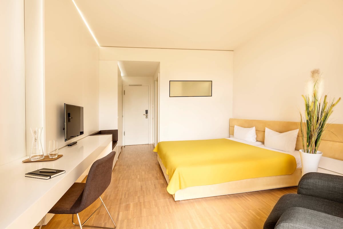 Quarto de hotel moderno com uma cama, uma mesa de trabalho com uma TV, uma cadeira e um vaso de planta.