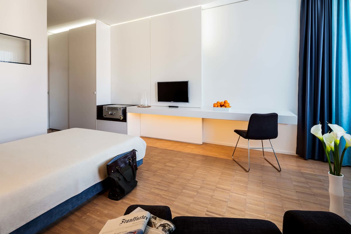 Quarto de hotel moderno com cama, secretária com televisão de tela plana, cadeira, vaso com flores e bolsa no chão.
