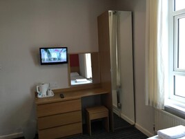 Quarto de hotel com cama, televisão na parede, escrivaninha de madeira com cadeira e janela com cortinas.