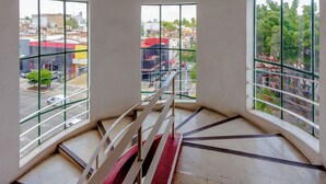 Canto de um quarto com escada que leva a um andar superior, grandes janelas com grades de metal e vista para um horizonte urbano com edifícios e árvores.