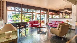 Um hall com um piano, sofás vermelhos e bege, uma mesa de centro e grandes janelas com vista para o exterior.