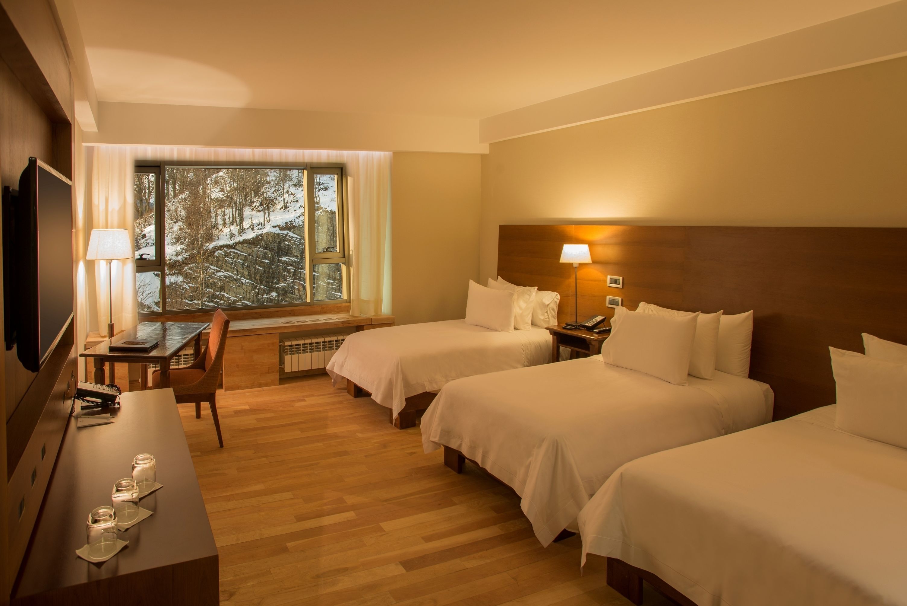 Quarto de hotel com duas camas, uma escrivaninha, uma cadeira, uma televisão e uma janela com vista para uma paisagem nevada.