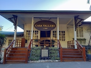 Um edifício com uma placa que diz "CASA VALLEJO c 1909" na entrada, com escadas de madeira e varanda coberta.