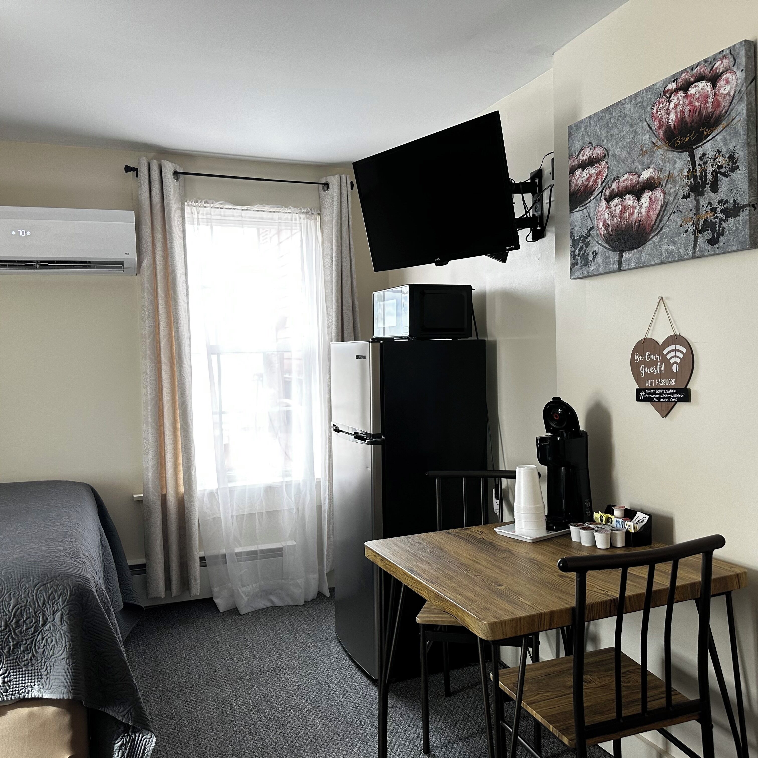 Quarto de hotel com cama, escrivaninha com cadeira, televisão de tela plana na parede, cafeteira e uma obra de arte emoldurada.