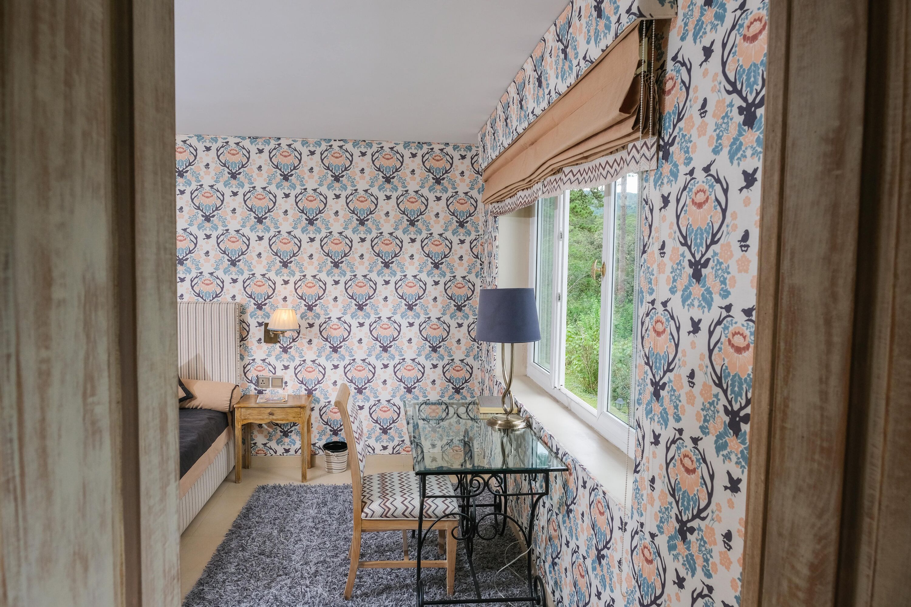 Quarto pequeno e acolhedor com papel de parede estampado, uma cama, uma escrivaninha com luminária e uma janela com vista para o verde.