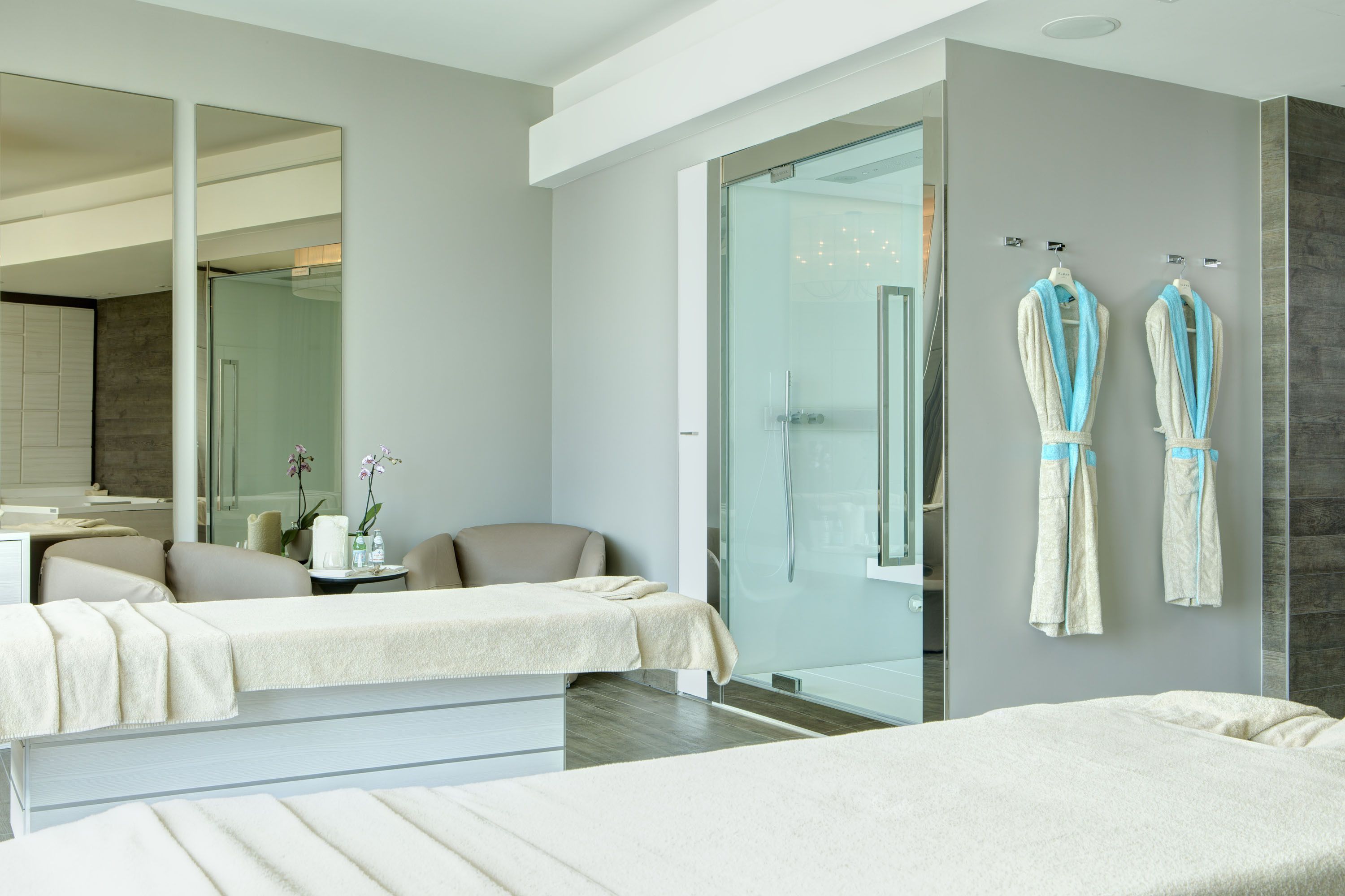 Quarto de hotel moderno com uma cama grande, uma área de estar separada, box de vidro e dois roupões pendurados na parede.