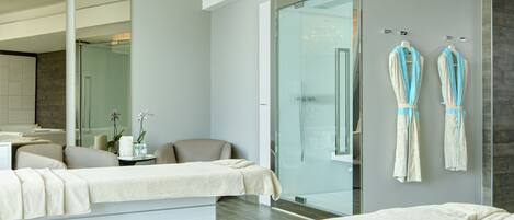 Quarto de hotel moderno com uma cama grande, uma área de estar separada, box de vidro e dois roupões pendurados na parede.