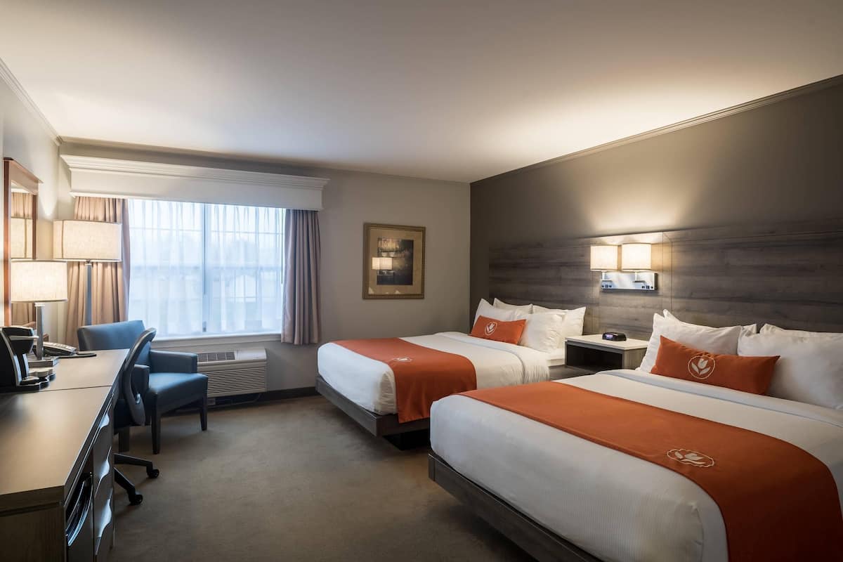Quarto de hotel com duas camas, uma escrivaninha, uma cadeira, um abajur, um quadro na parede e uma janela com cortinas.