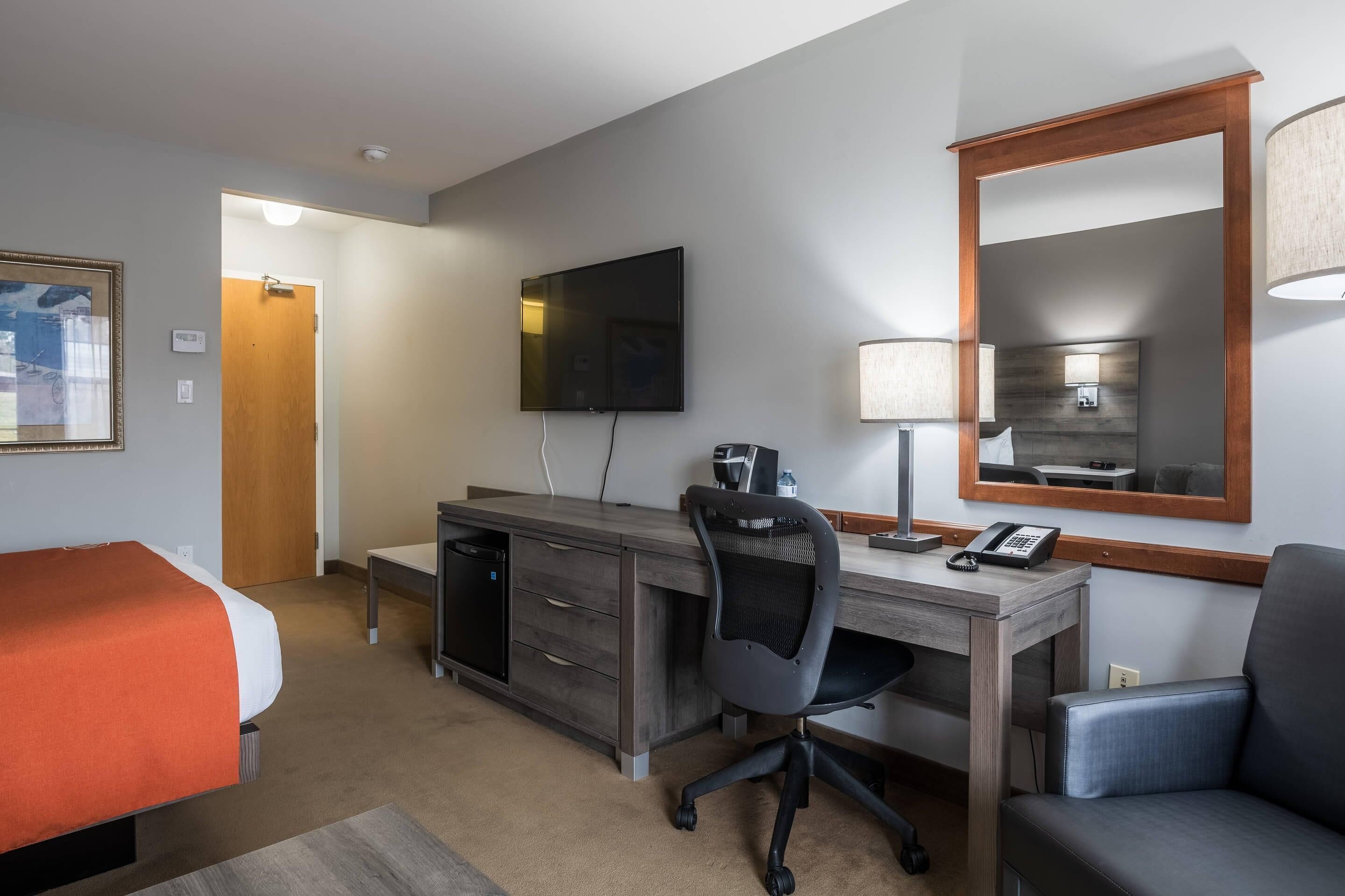 Quarto de hotel com cama, escrivaninha, cadeira, televisão e espelho.