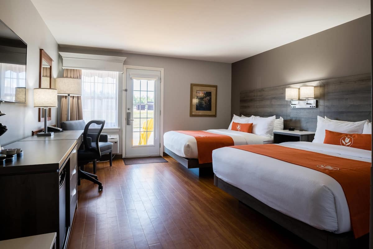 Quarto de hotel com duas camas, uma escrivaninha, uma cadeira, um abajur, um quadro e uma janela.