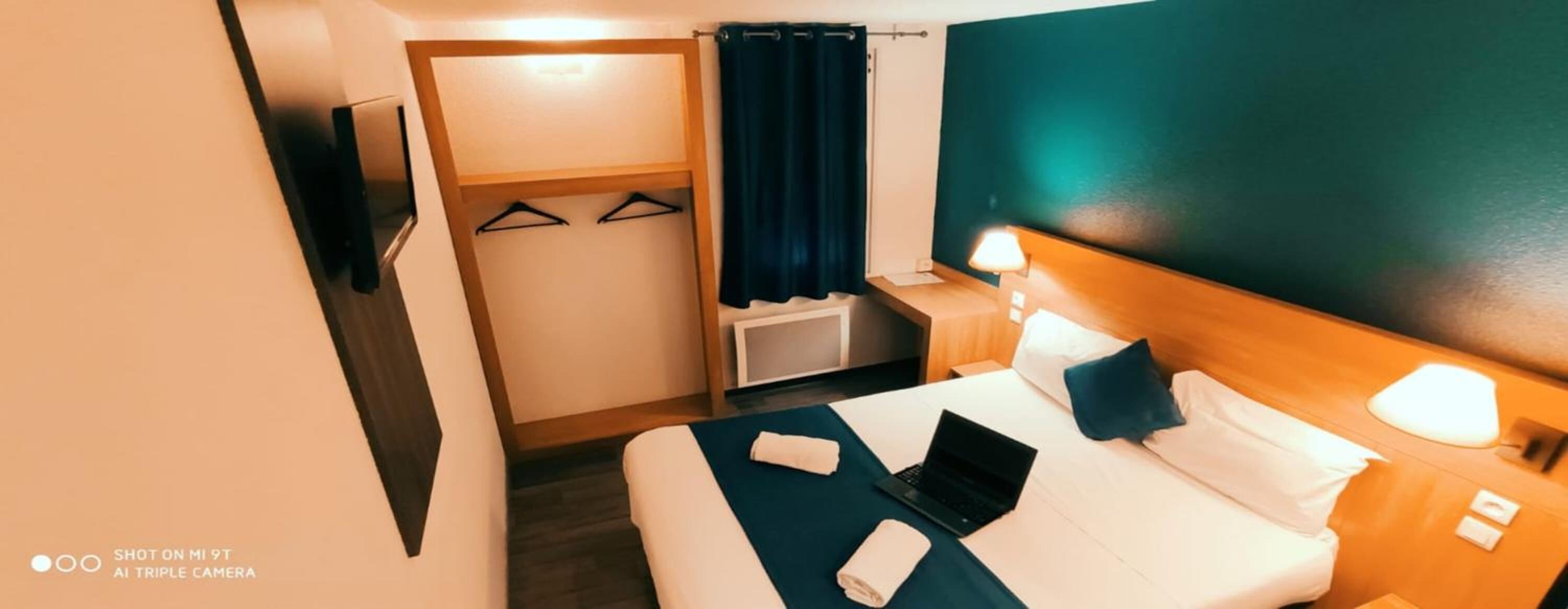 Quarto de hotel com cama, luminárias de cabeceira, televisão, cabideiro e área de chuveiro.