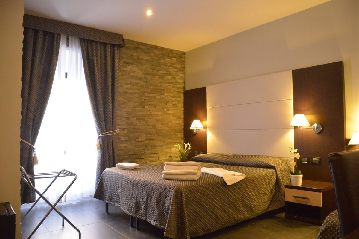 Quarto de hotel com duas camas, televisão fixada na parede, mesa de cabeceira com abajur e janela com cortinas.