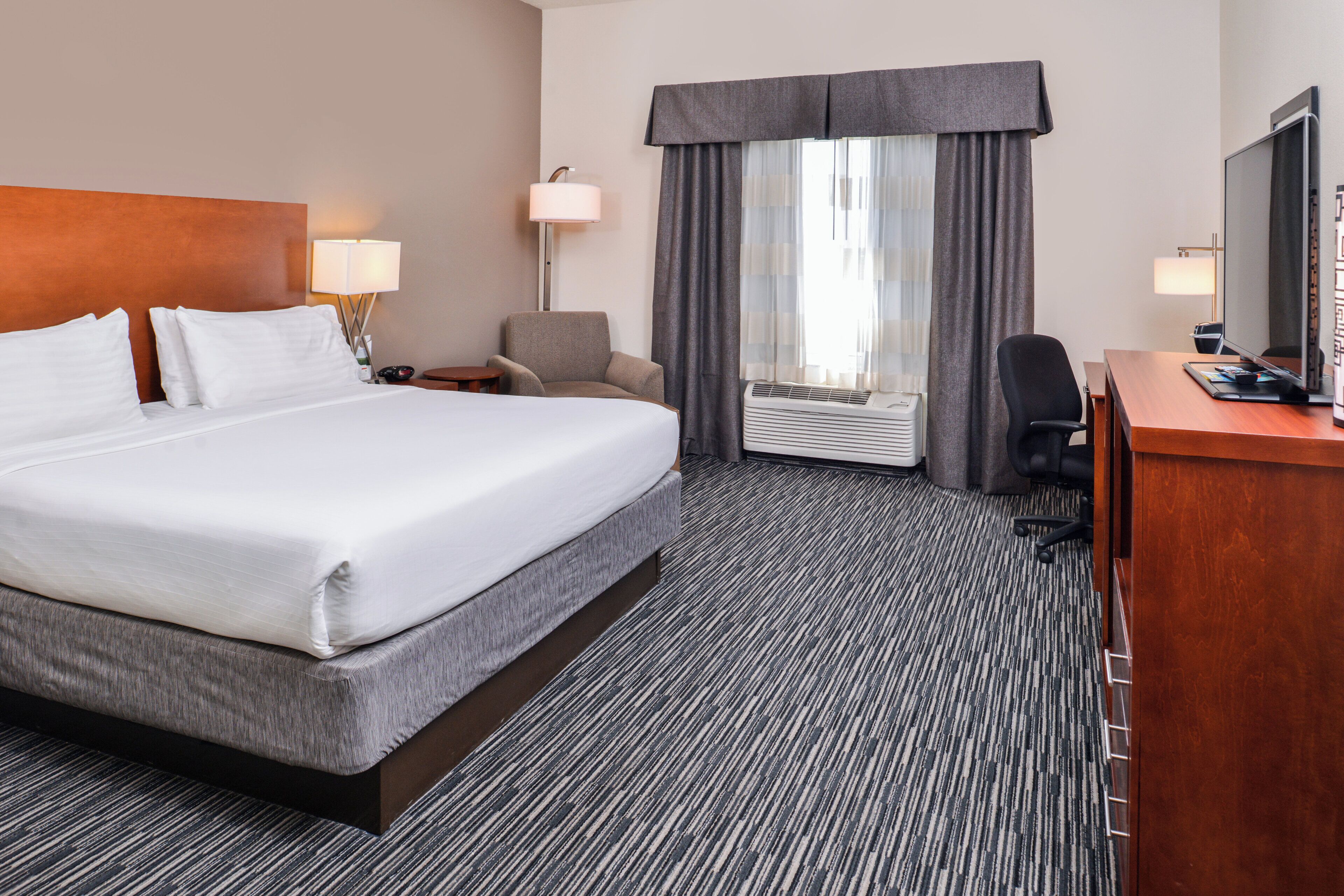 Quarto de hotel com uma cama grande, uma escrivaninha, uma cadeira, um abajur e uma televisão.