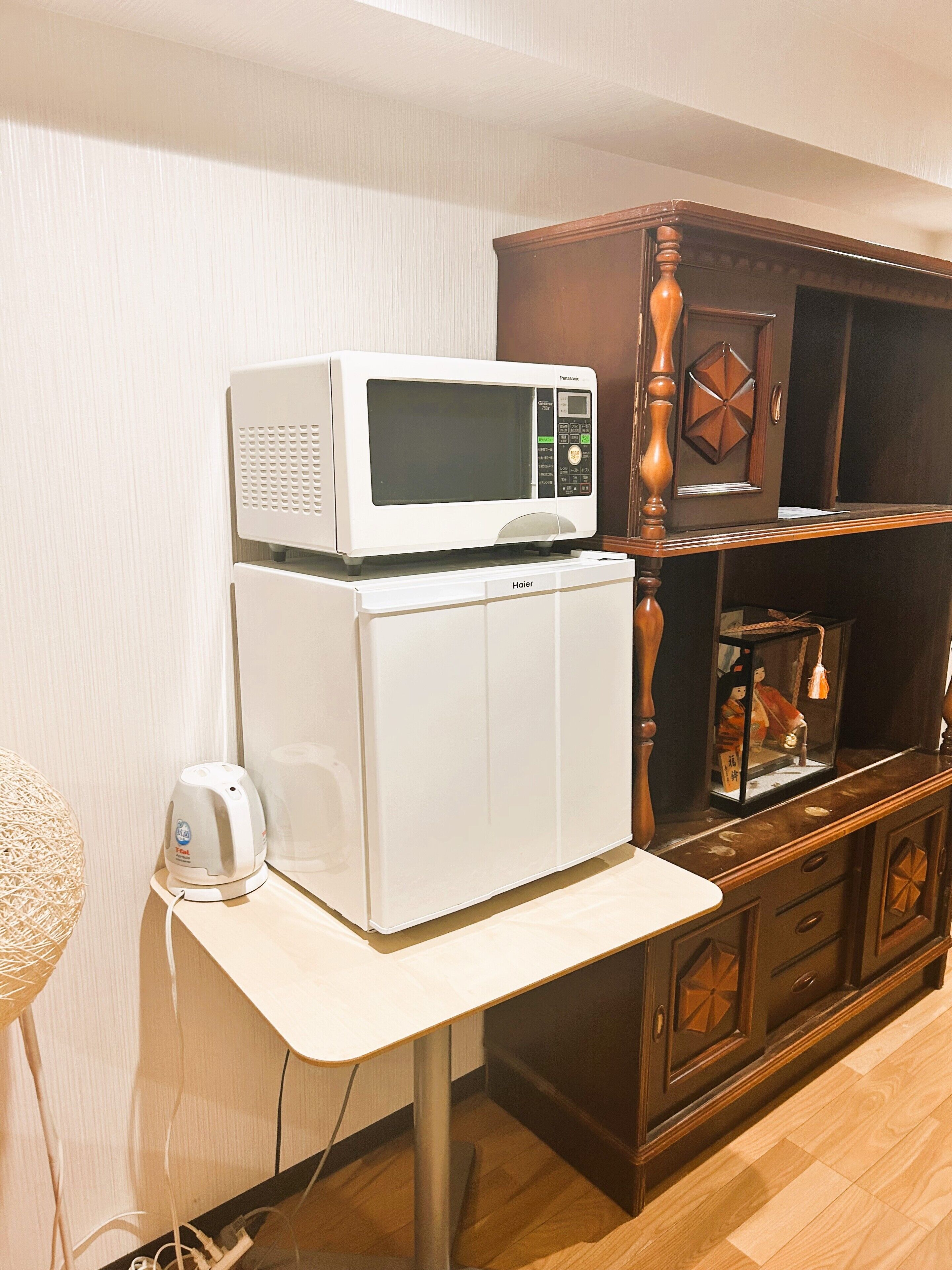 Uma cozinha compacta com micro-ondas sobre a geladeira, uma mesa pequena com uma chaleira e um armário de madeira com um objeto decorativo.