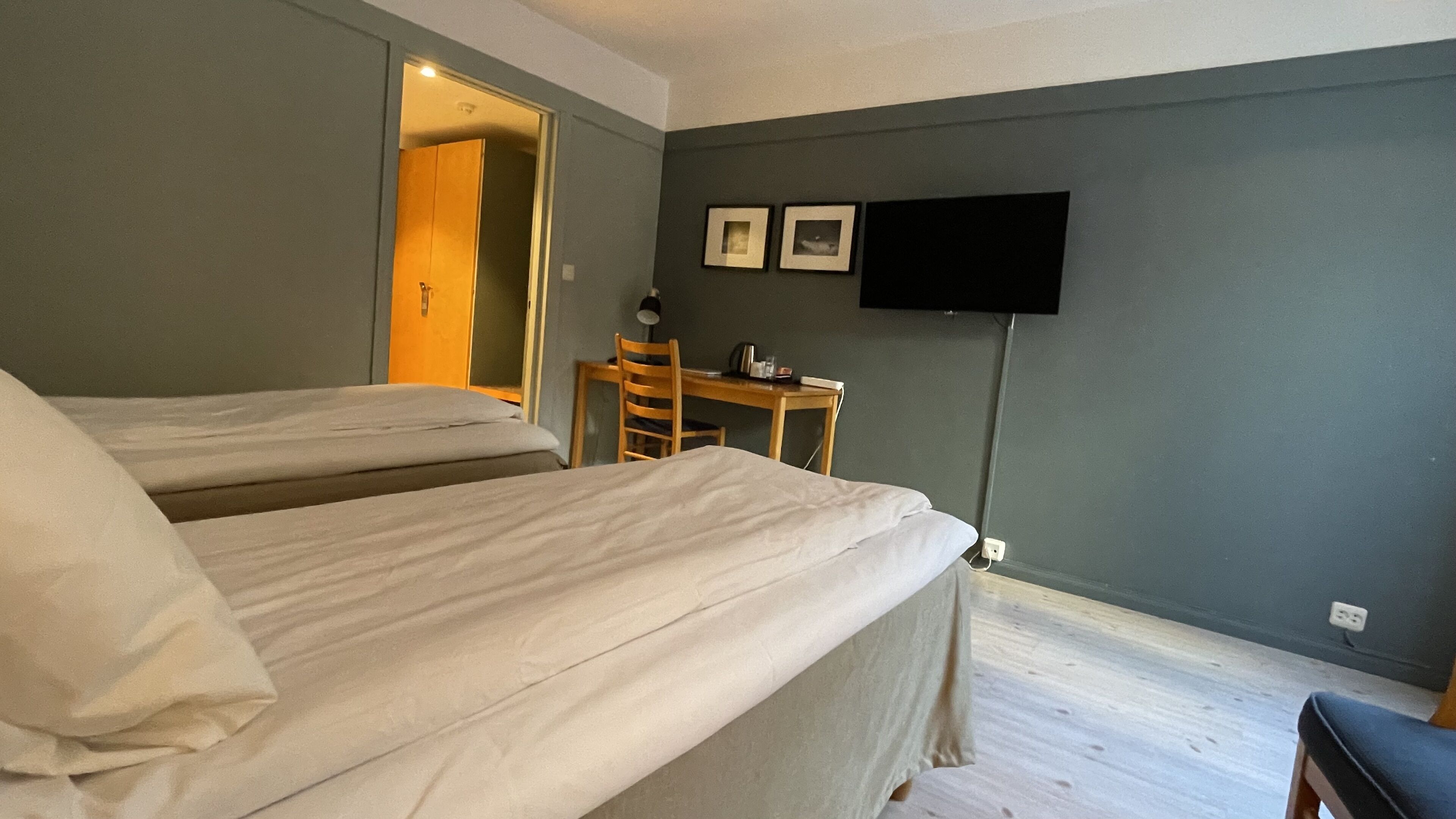 Quarto de hotel com duas camas, uma escrivaninha, uma cadeira e televisão de tela plana fixada na parede.