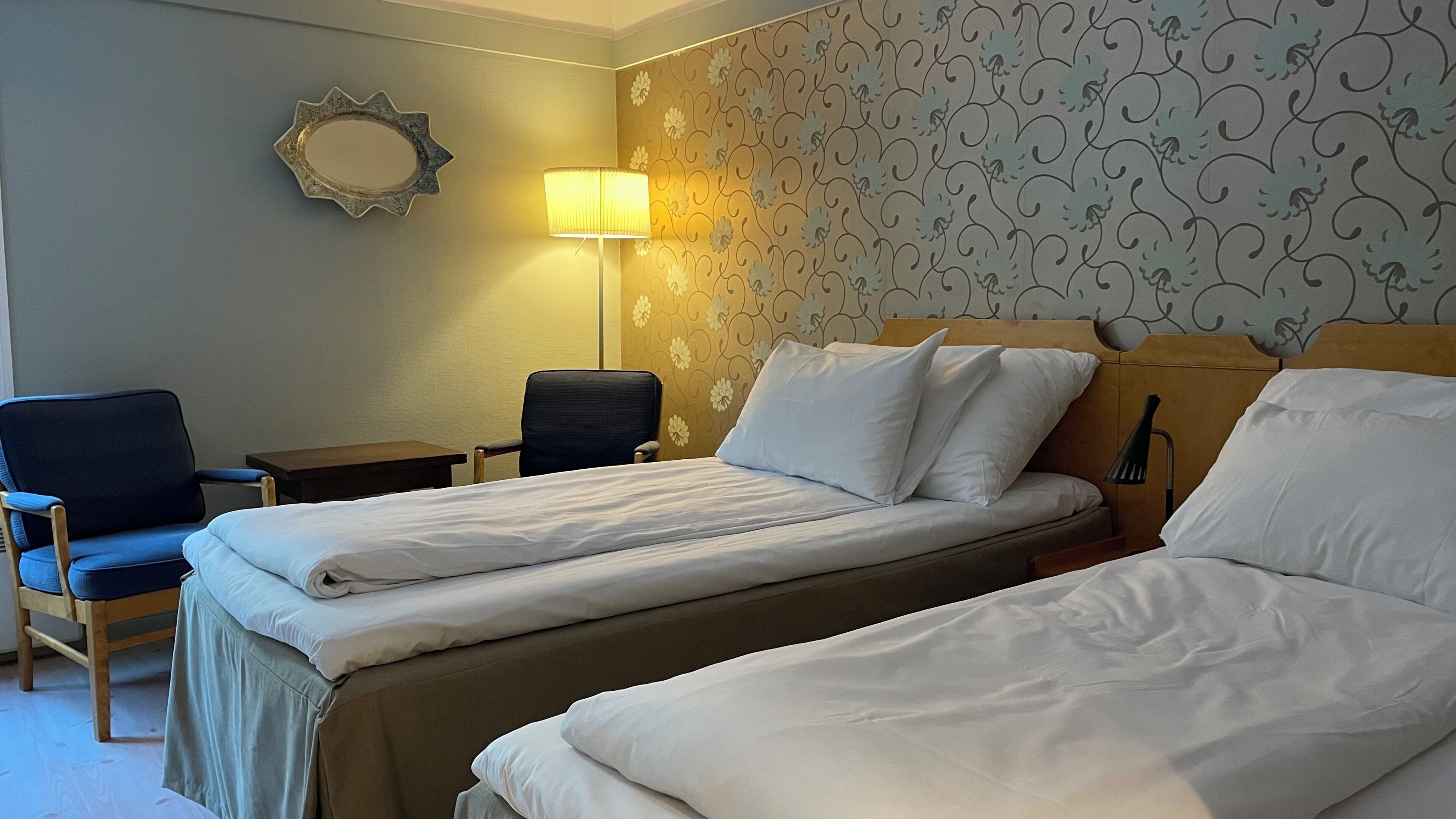 Quarto de hotel com duas camas, uma mesa, uma cadeira, uma luminária e um espelho decorativo.