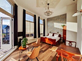 Quarto de hotel moderno com uma cama grande, uma mesa de jantar e cadeiras. Há janelas com cortinas, um candelabro e uma planta em vaso.