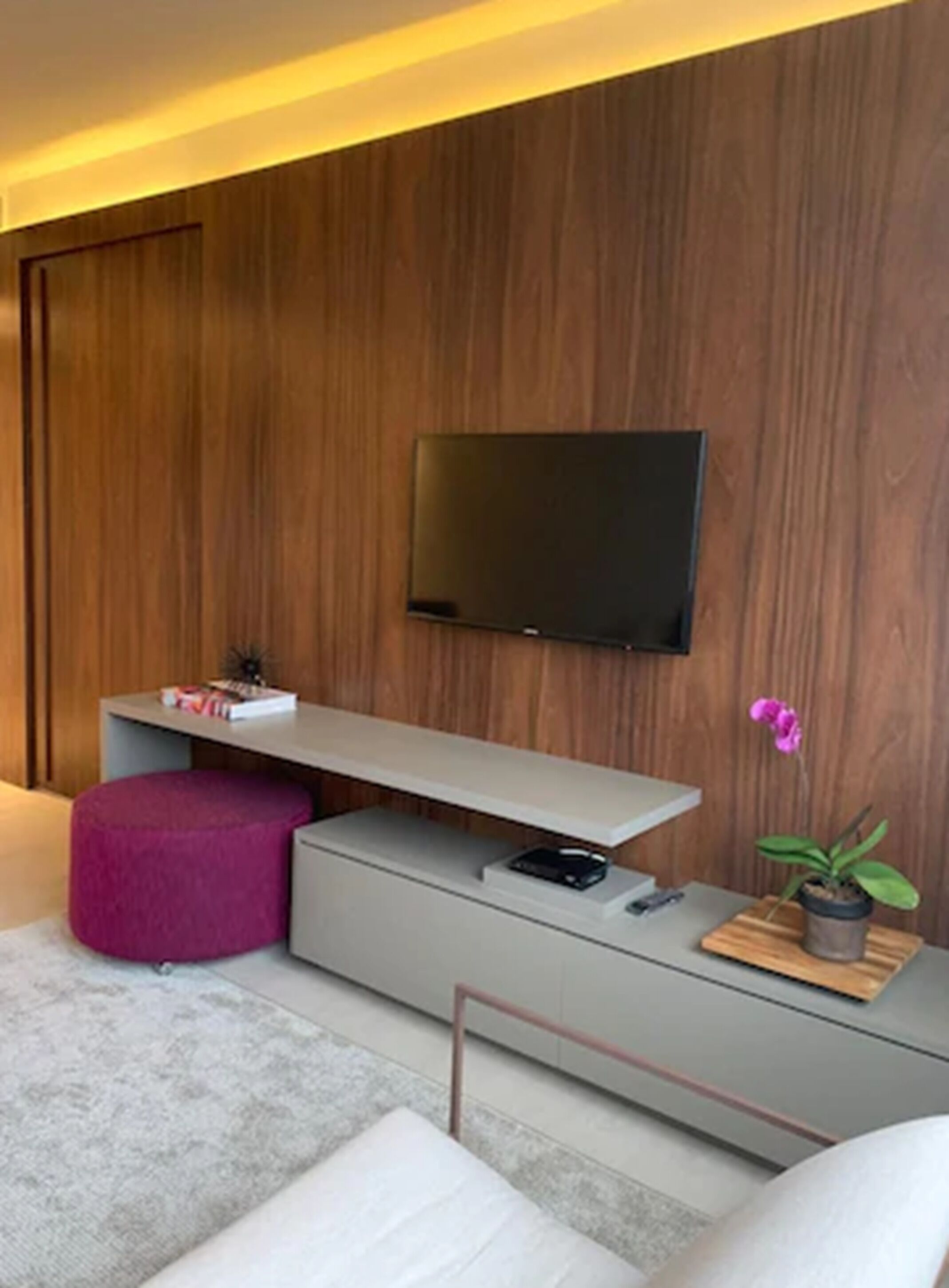 Uma sala de estar moderna com uma TV de tela plana montada em uma parede de madeira, um sofá cinza, um banquinho rosa pequeno e uma mesa lateral de madeira com uma planta em vaso.