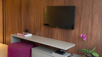 Uma sala de estar moderna com uma TV de tela plana montada em uma parede de madeira, um sofá cinza, um banquinho rosa pequeno e uma mesa lateral de madeira com uma planta em vaso.