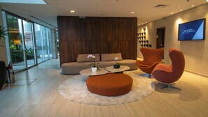 Um lobby moderno com sofá, um banquinho redondo, duas poltronas, uma tela de TV e uma parede de madeira.