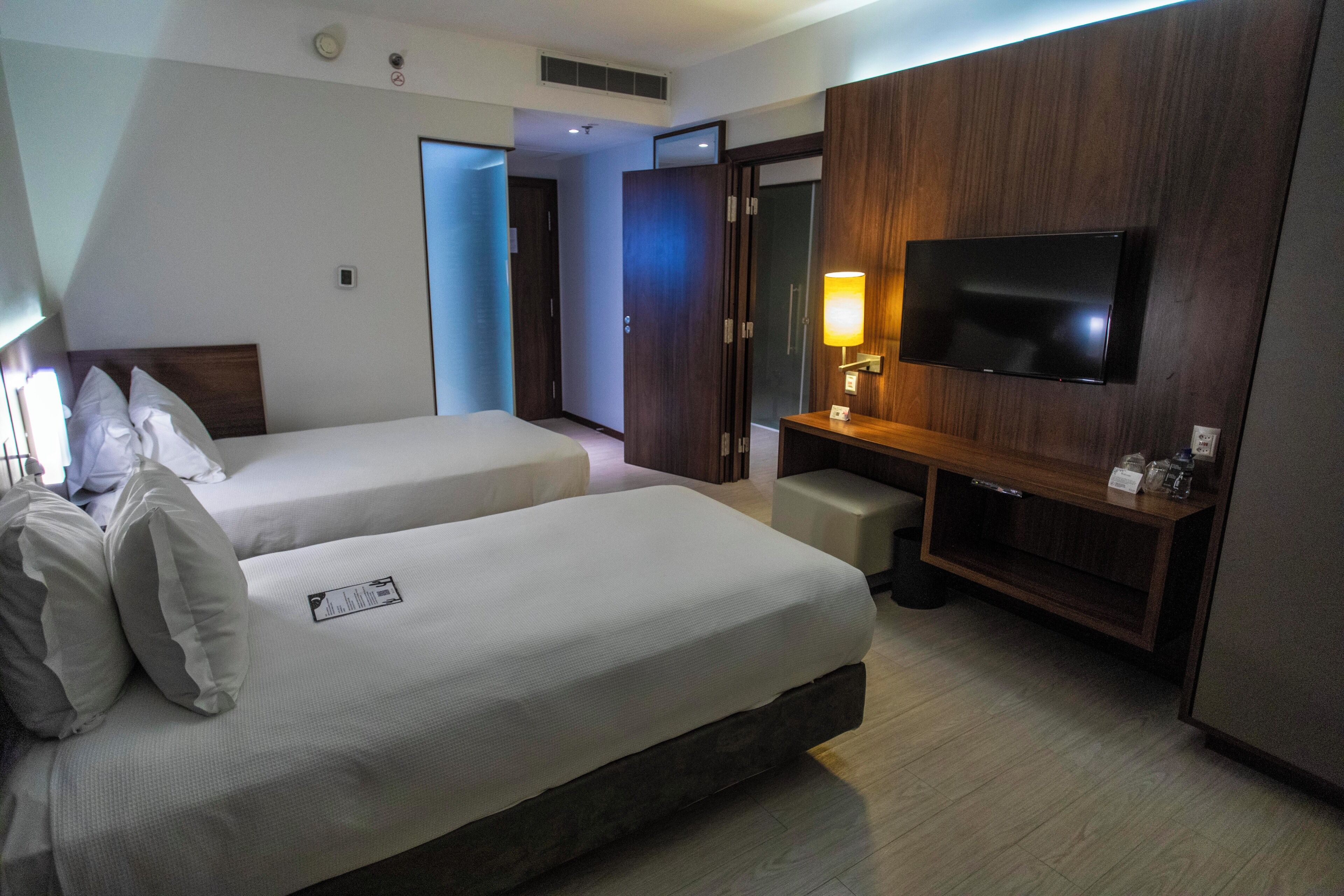 Quarto de hotel com duas camas, uma escrivaninha com televisão e um guarda-roupa de madeira.