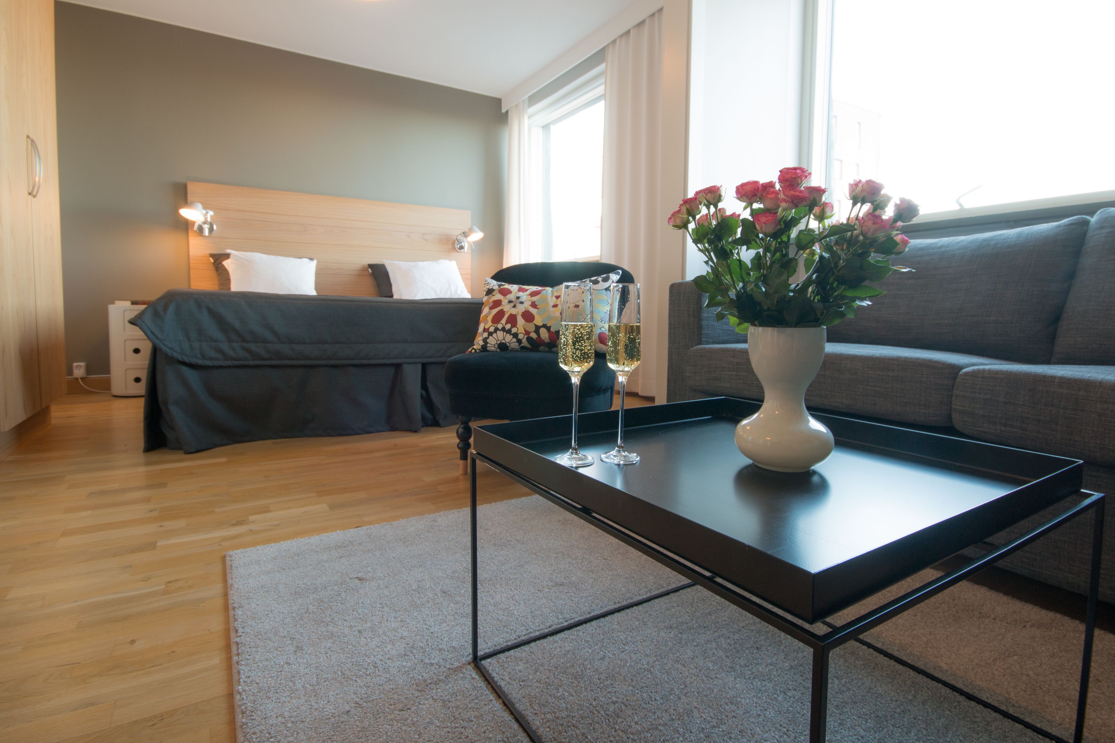 Quarto de hotel com cama, sofá, mesa de centro com um vaso de flores e duas taças de champanhe.