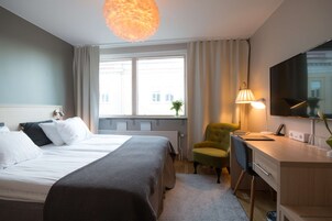 Quarto de hotel com cama, escrivaninha, cadeira, luminária, televisão e janela com cortinas.