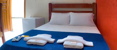 Quarto de hotel com uma cama, um cobre-leito azul, almofadas brancas e uma cabeceira de madeira. Há toalhas na cama e um controle remoto ao lado.