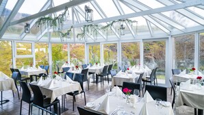 Um restaurante com teto de vidro, mesas com toalhas brancas e cadeiras pretas, e plantas penduradas.