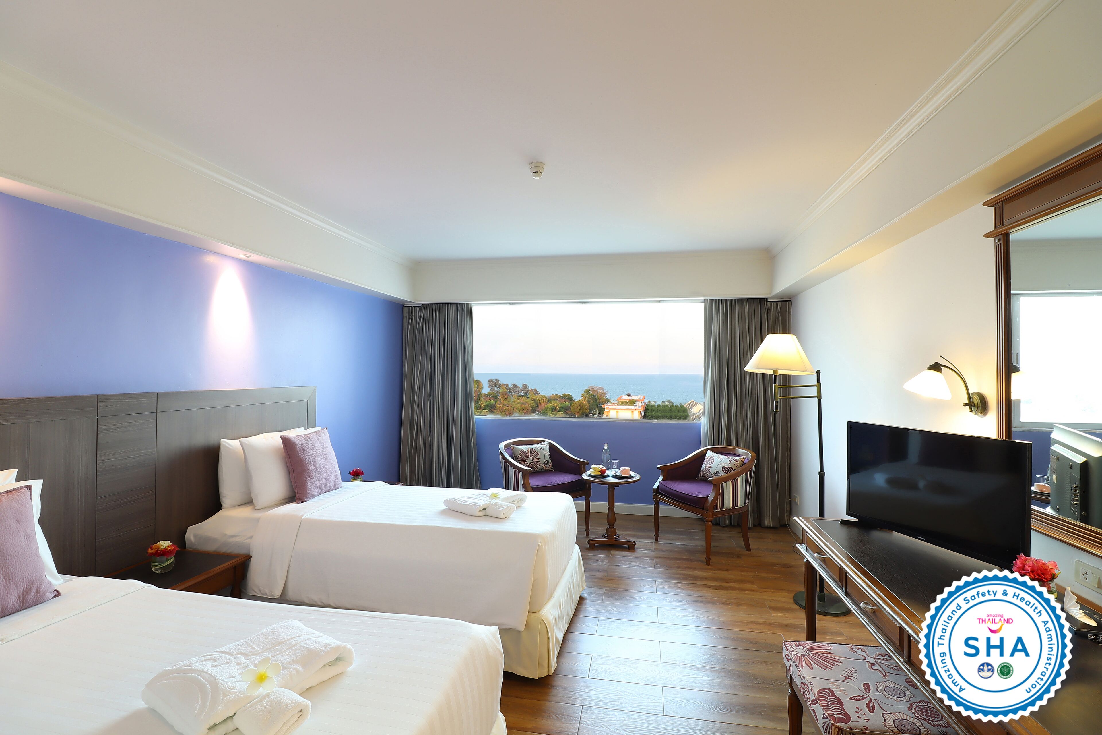 Quarto de hotel com duas camas, televisão, uma mesinha com abajur e varanda com vista.