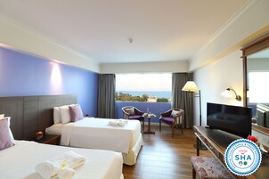Quarto de hotel com duas camas, televisão, uma mesinha com abajur e varanda com vista.