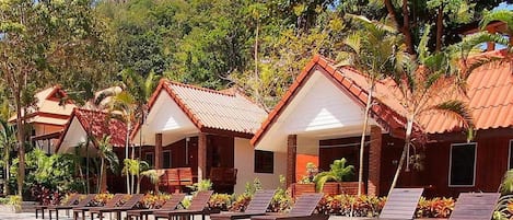 Área à beira da piscina com espreguiçadeiras e piscina, cercada por vegetação tropical e edifícios com telhados vermelhos.